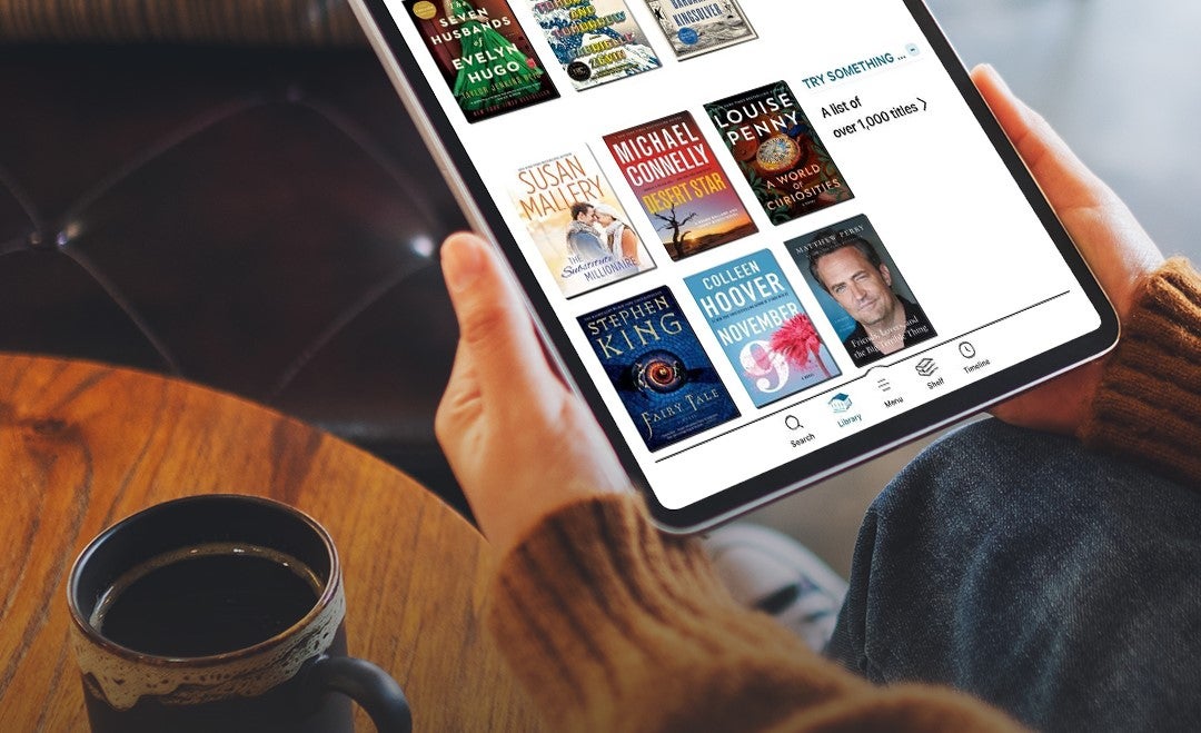 Person holds an e-reader with several book titles visible; a small table and mug are in the background